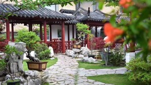 Um jardim chinês tradicional, com caminhos de pedra, estruturas de madeira vermelha e diversas plantas.