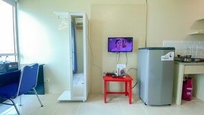 Uma pequena cozinha compacta com geladeira, micro-ondas e pia. Uma televisão está fixada na parede, exibindo um programa. Uma cadeira e uma mesa pequena são visíveis no espaço.