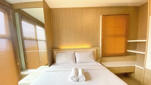 Cama bem arrumada com lençóis brancos e duas toalhas enroladas, cabeceira de madeira e uma janela com persianas.