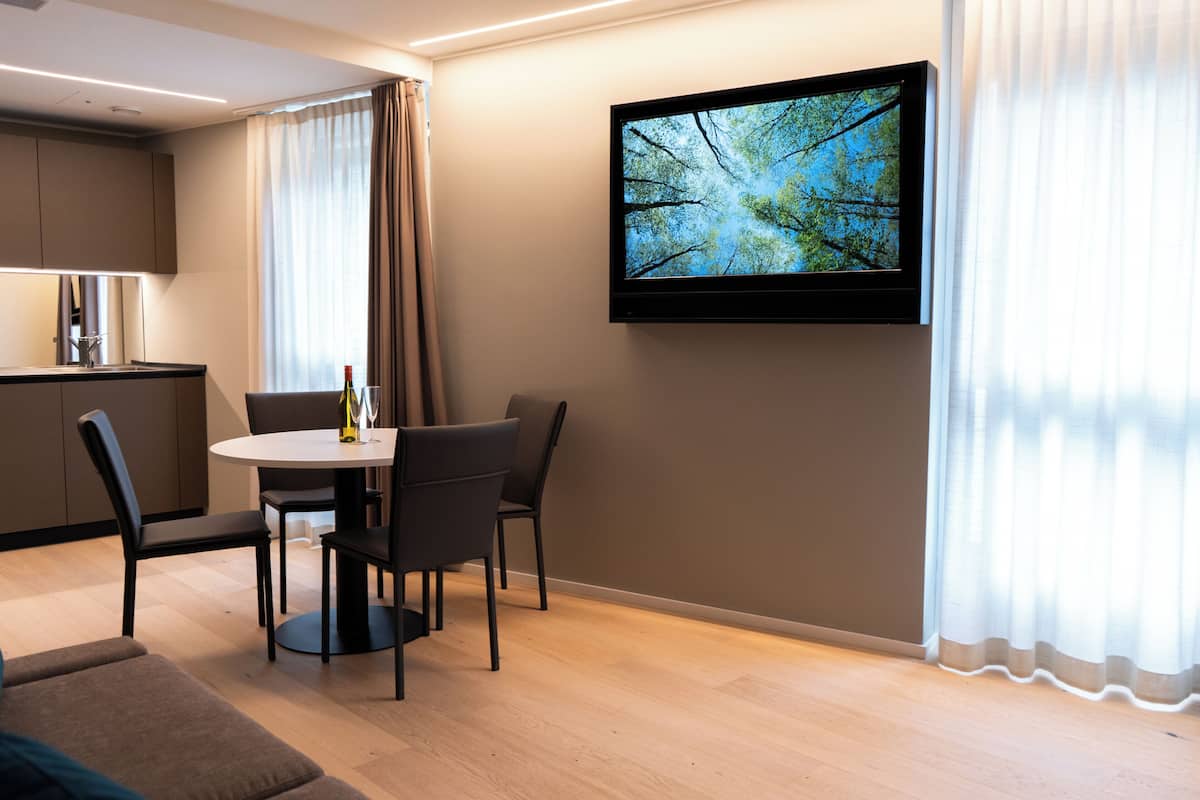Quarto de hotel moderno com área de refeições, televisão de tela plana na parede e vista para árvores pela janela.