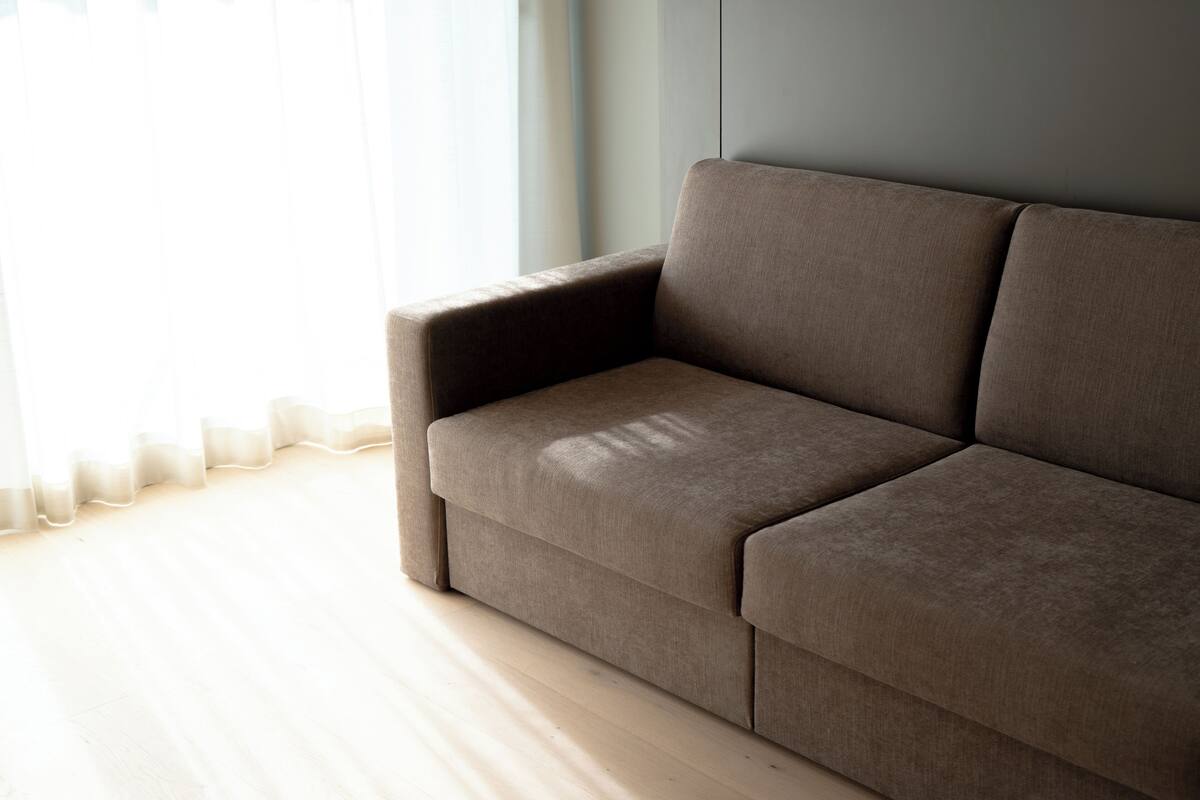Uma sala de estar moderna com um sofá marrom, piso de madeira clara e cortinas leves.