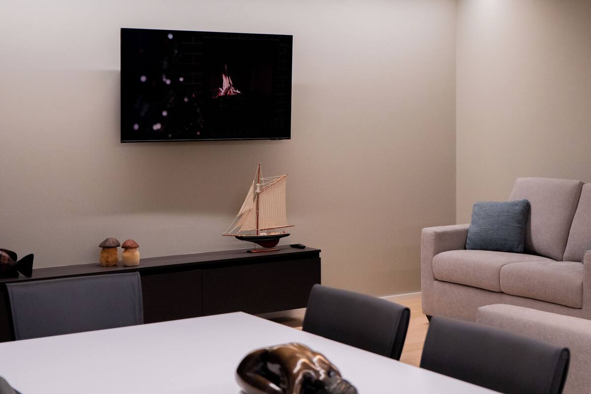 Uma sala de estar moderna com uma TV de tela plana na parede, um modelo de veleiro pequeno em cima de um console escuro e um sofá bege com uma almofada azul.