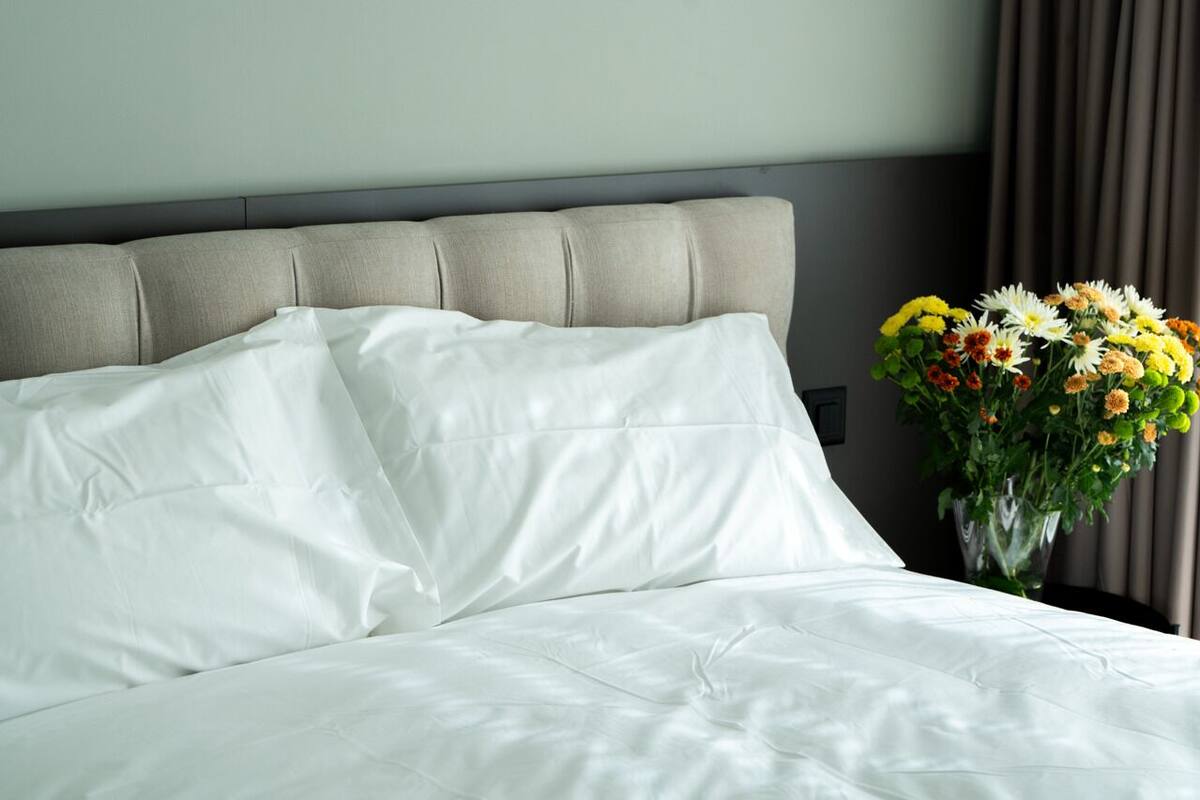 Cama bem arrumada com roupa de cama branca e cabeceira estofada cinza. Um vaso com flores amarelas e laranjas está ao lado da cama.