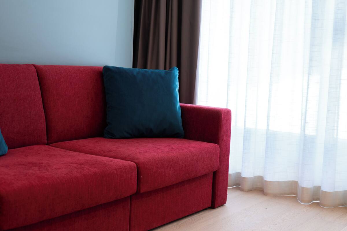 Um sofá vermelho com um travesseiro azul-esverdeado em um quarto com piso de madeira clara e cortinas translúcidas.