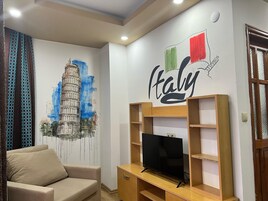 Uma sala de estar com um grande mural na parede retratando a Torre de Pisa, uma televisão sobre um suporte de madeira e um sofá.