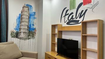 Uma sala de estar com um sofá bege, uma TV de tela plana sobre um suporte de madeira e um mural na parede da Torre de Pisa.