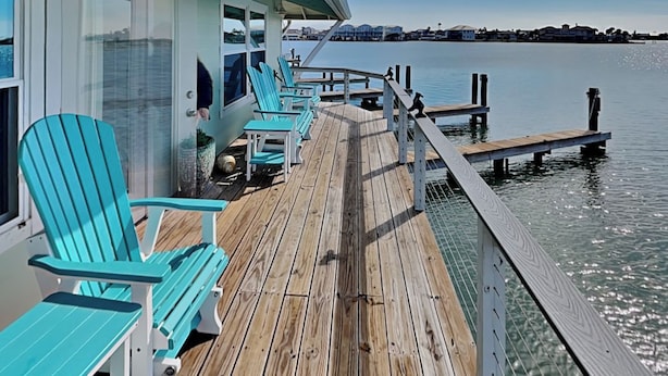 Um deck de madeira com cadeiras Adirondack turquesa, corrimão e vista para a água e edifícios ao longe.