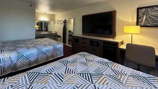 Quarto de hotel com duas camas, televisão, secretária com candeeiro, uma cadeira e casa de banho visível ao fundo.