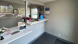 Uma área de recepção com divisória de vidro, um relógio, um aviso indicando a disponibilidade do serviço, um vaso de flores e um código QR em um balcão.