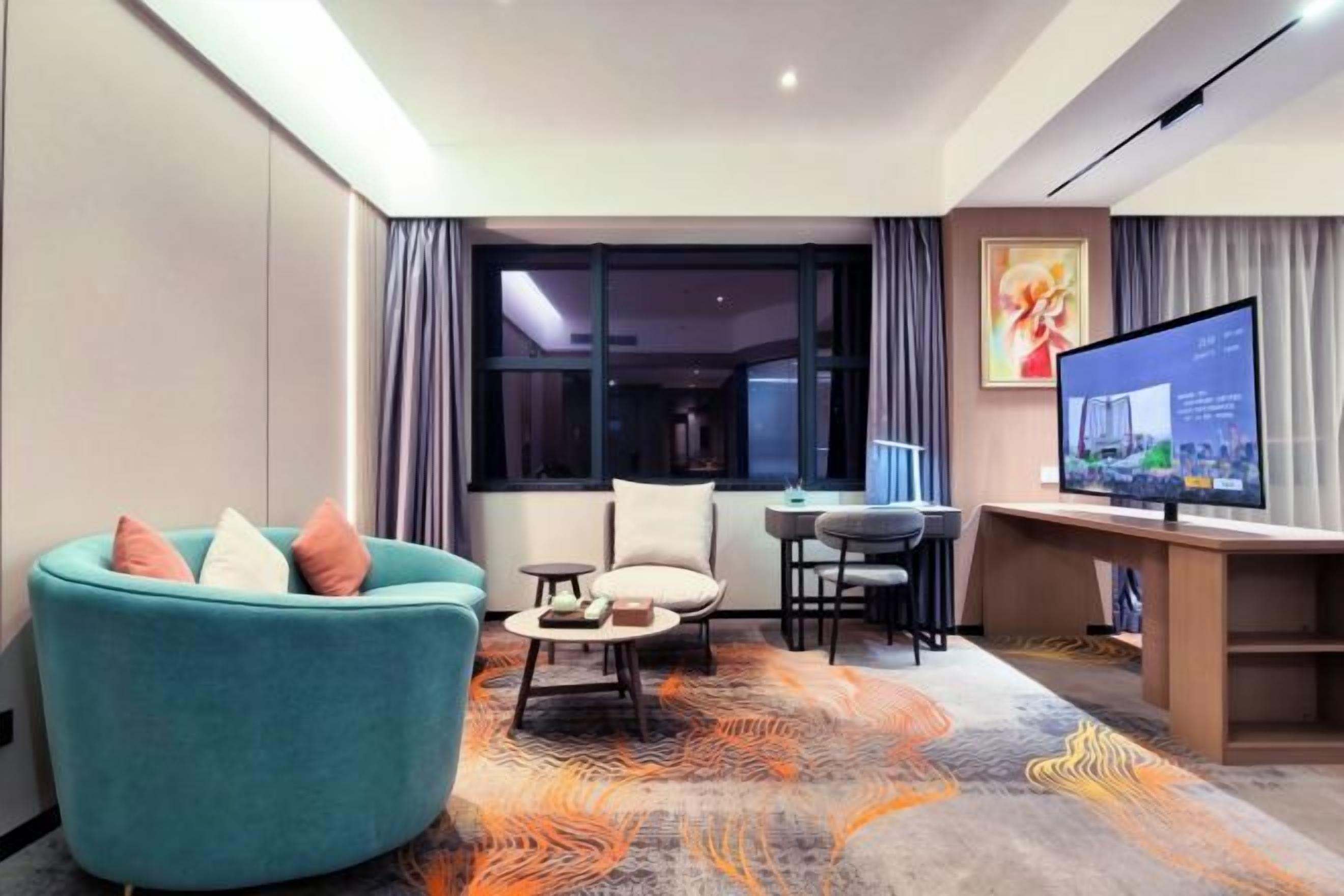 Quarto de hotel moderno com sofá verde-água curvo, mesa de centro redonda, televisão e área de refeições com mesa e cadeiras.
