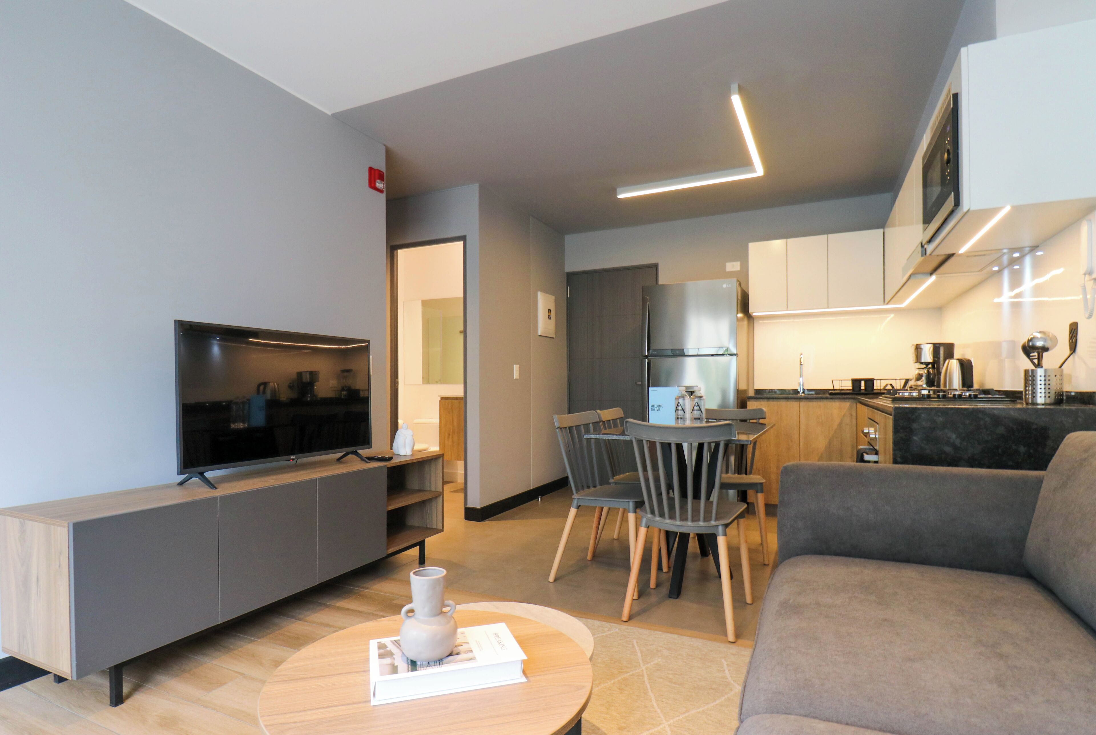 Uma sala de estar moderna com televisão de tela plana, sofá cinza, mesa de centro de madeira e um espaço de jantar com mesa e cadeiras.