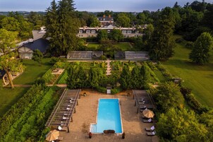 Uma piscina grande e retangular rodeada por espreguiçadeiras e guarda-sóis, com um jardim bem cuidado e uma casa grande ao fundo.