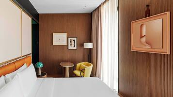 Quarto de hotel com uma cama grande, uma mesa redonda pequena, uma poltrona amarela e uma obra de arte pendurada na parede.