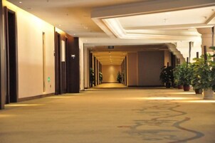 Corredor de hotel com portas em ambos os lados, piso coberto de carpete e plantas em vasos ao longo da parede direita.