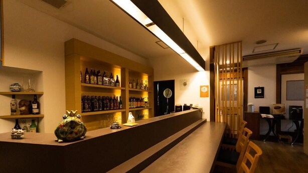 Uma área de bar com um balcão comprido, prateleiras com garrafas e uma figura decorativa de um sapo.