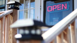 Uma cerca de madeira com uma câmera de segurança e um letreiro de neon 'OPEN' ao fundo.