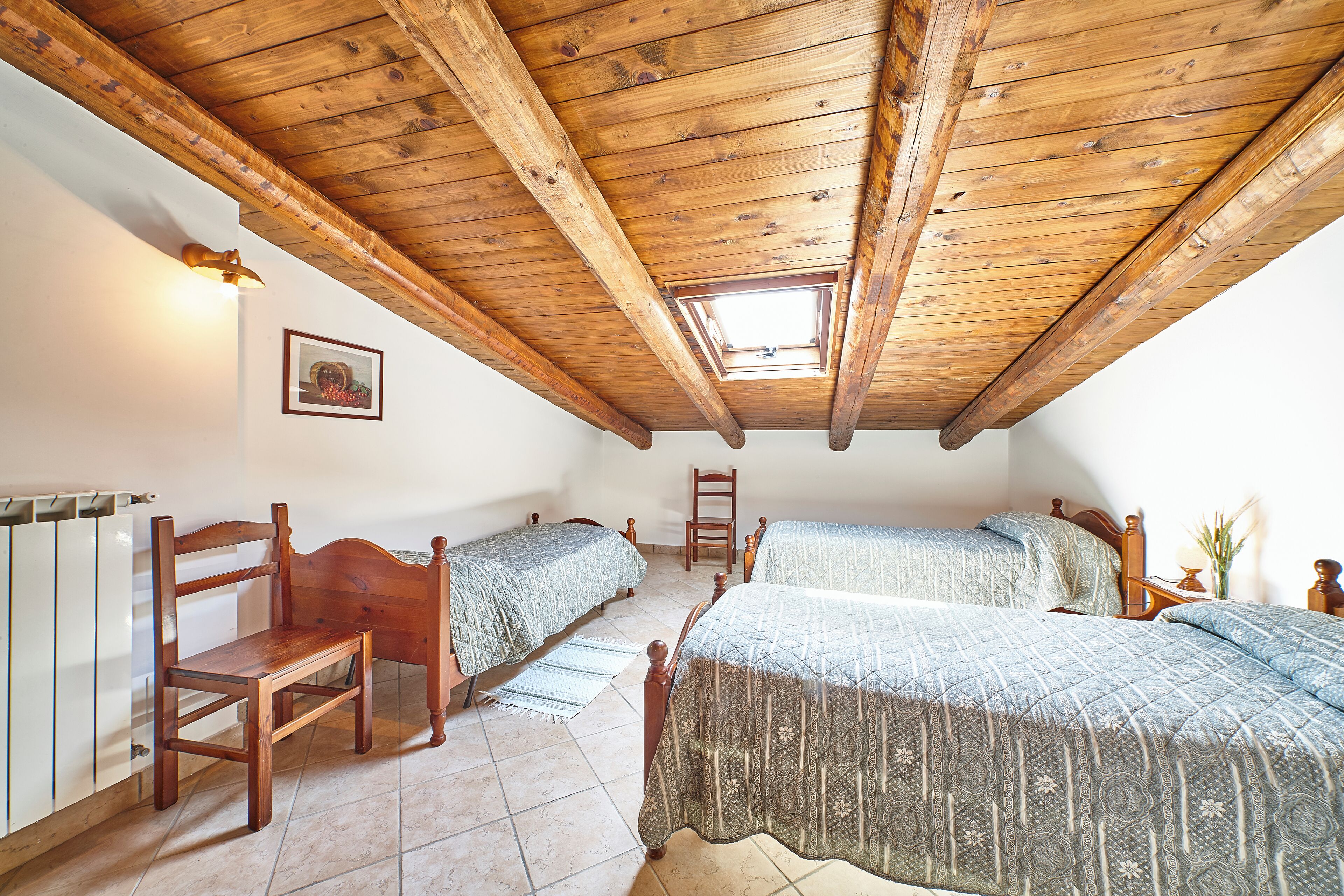 Quarto com duas camas, uma cadeira de madeira, um radiador e um claraboia.