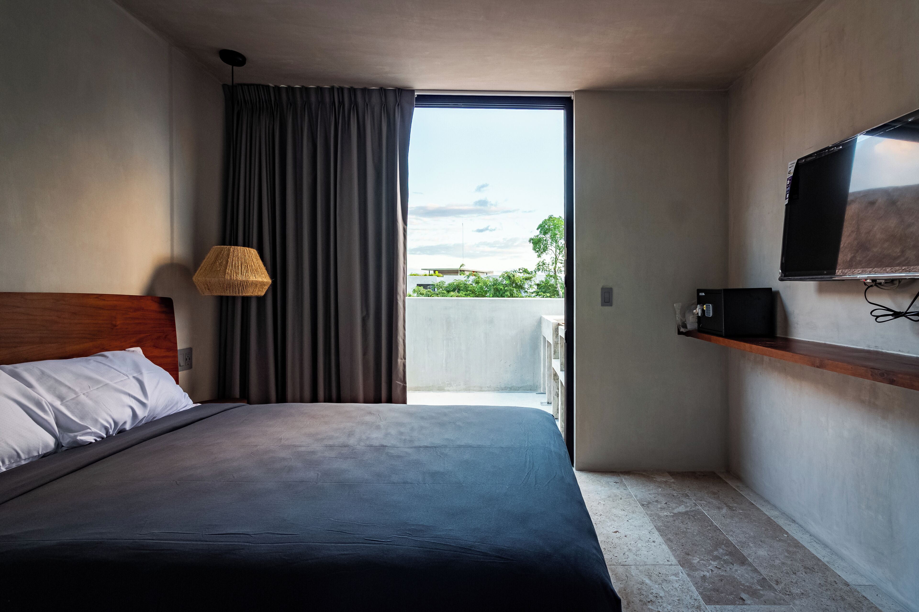 Quarto com cama, televisão, pequena estante, janela com cortinas e vista para o exterior.