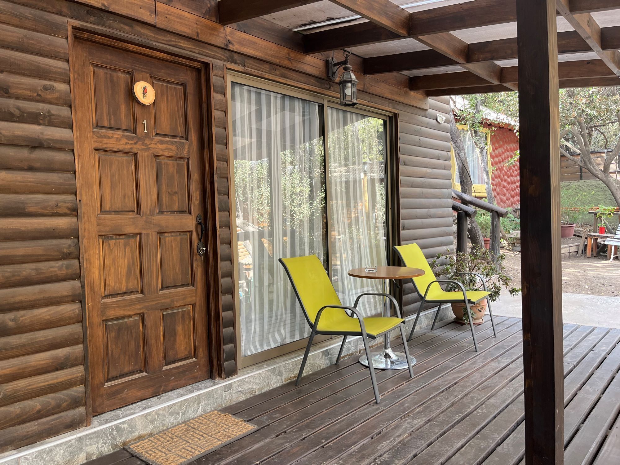 Uma cabana de toras de madeira com uma grande porta de madeira, uma porta de vidro deslizante e uma varanda com duas cadeiras amarelas e uma mesa redonda pequena.