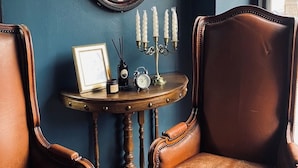 Um quarto com duas poltronas de couro, um espelho redondo, uma mesa pequena com moldura de quadro, velas e uma luminária na parede.