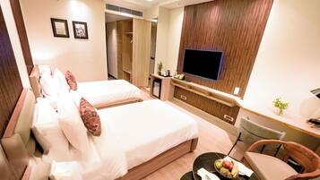 Quarto de hotel com duas camas, uma televisão, uma escrivaninha com cadeira e uma mesinha com fruta.