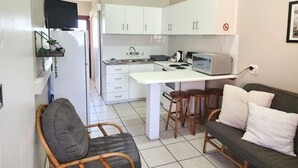 Cozinha compacta com armários brancos, micro-ondas e pia. Há uma pequena área de refeições com bancos e um sofá.