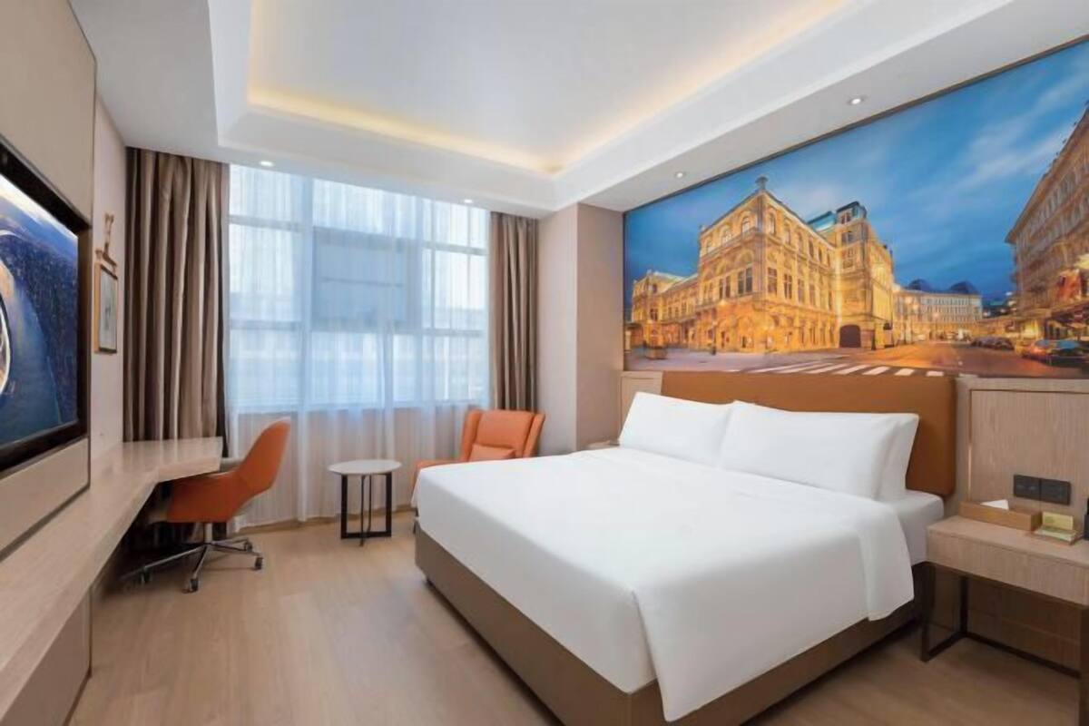 Quarto de hotel com uma cama grande, uma mesa de trabalho, uma poltrona laranja e um quadro com vista para a cidade na parede.
