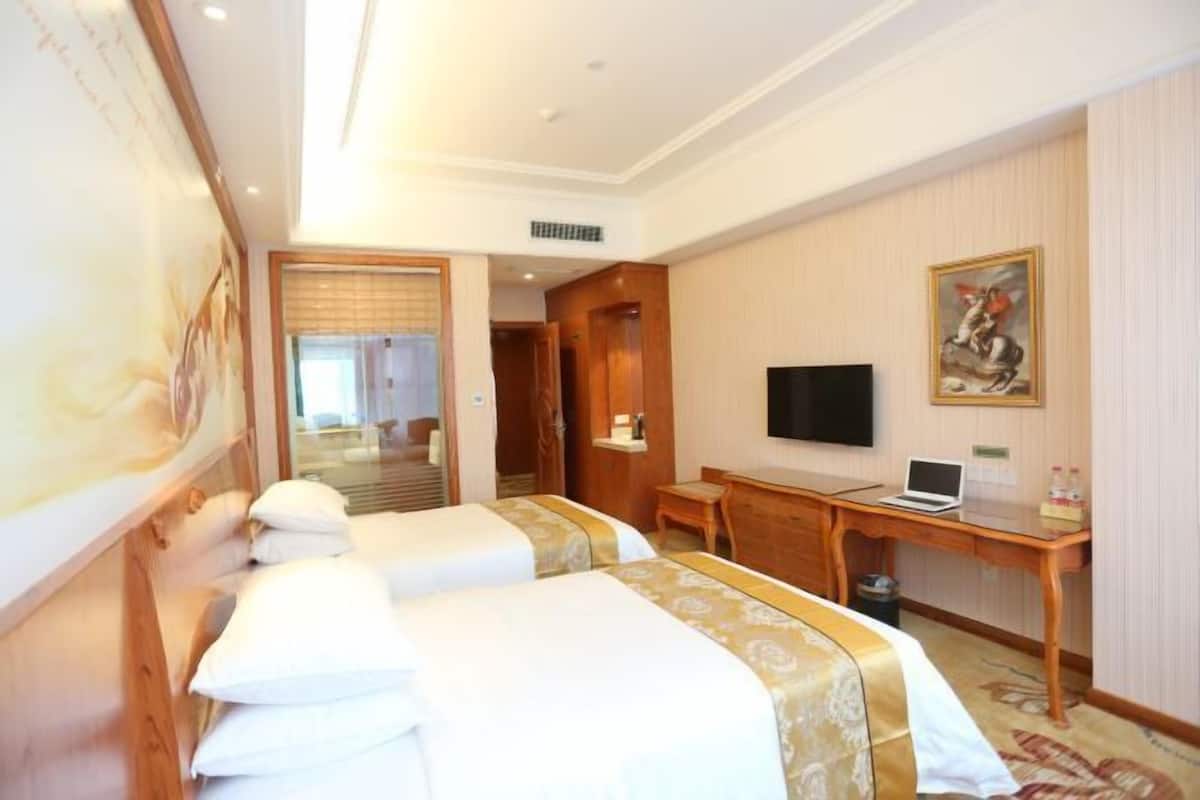 Quarto de hotel com duas camas, uma escrivaninha, televisão e um quadro na parede.