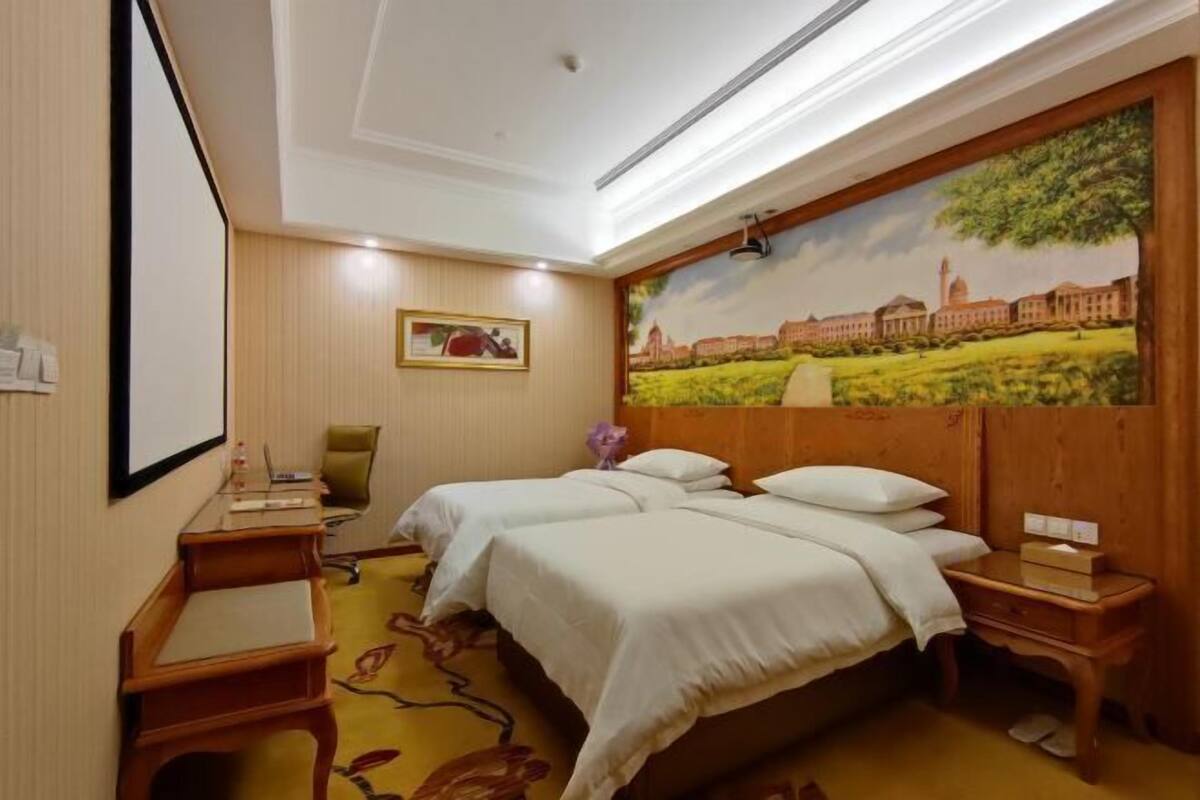 Quarto de hotel com duas camas, uma escrivaninha, uma cadeira e um quadro de uma paisagem.