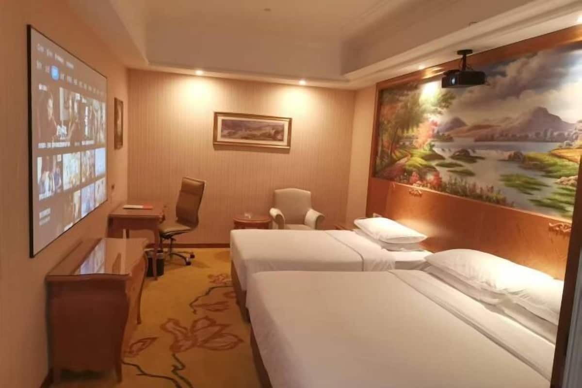 Quarto de hotel com duas camas, uma escrivaninha, uma cadeira, uma tela de projetor e um mural na parede.