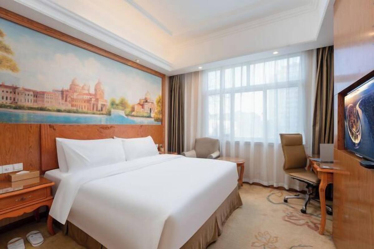 Quarto de hotel com uma cama grande, uma escrivaninha, uma cadeira, uma televisão e um quadro na parede.