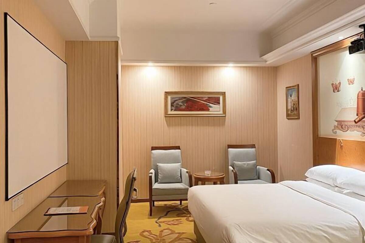Quarto de hotel com cama, duas poltronas, uma mesa pequena, uma televisão e um quadro na parede.