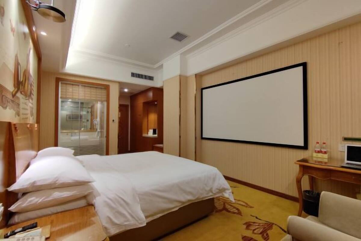 Quarto de hotel com cama, secretária com computador, cadeira, tela de projeção e janela com persianas.