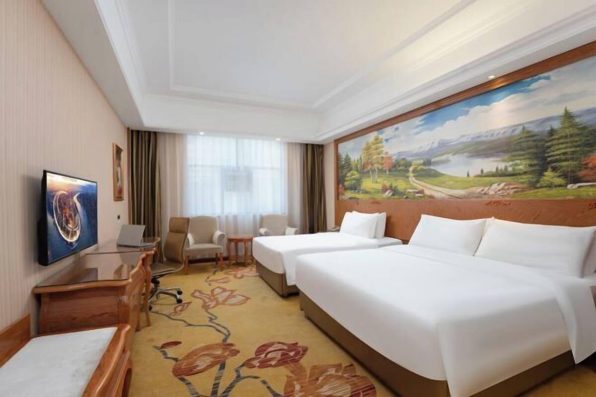 Quarto de hotel com duas camas, uma mesa de trabalho com um laptop, uma cadeira e um quadro na parede.