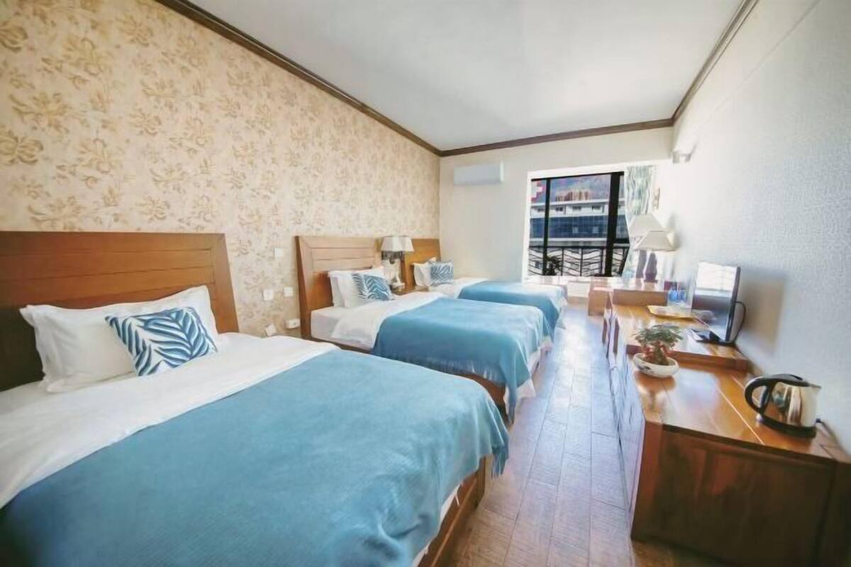 Quarto de hotel com três camas, cabeceira de madeira, escrivaninha com abajur e janela com vista para edifícios.
