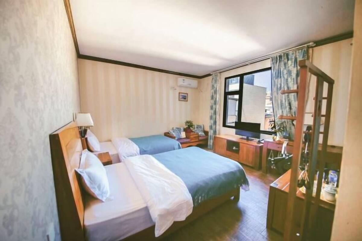 Quarto de hotel com duas camas, uma escrivaninha, uma cadeira, televisão e janela com cortinas.