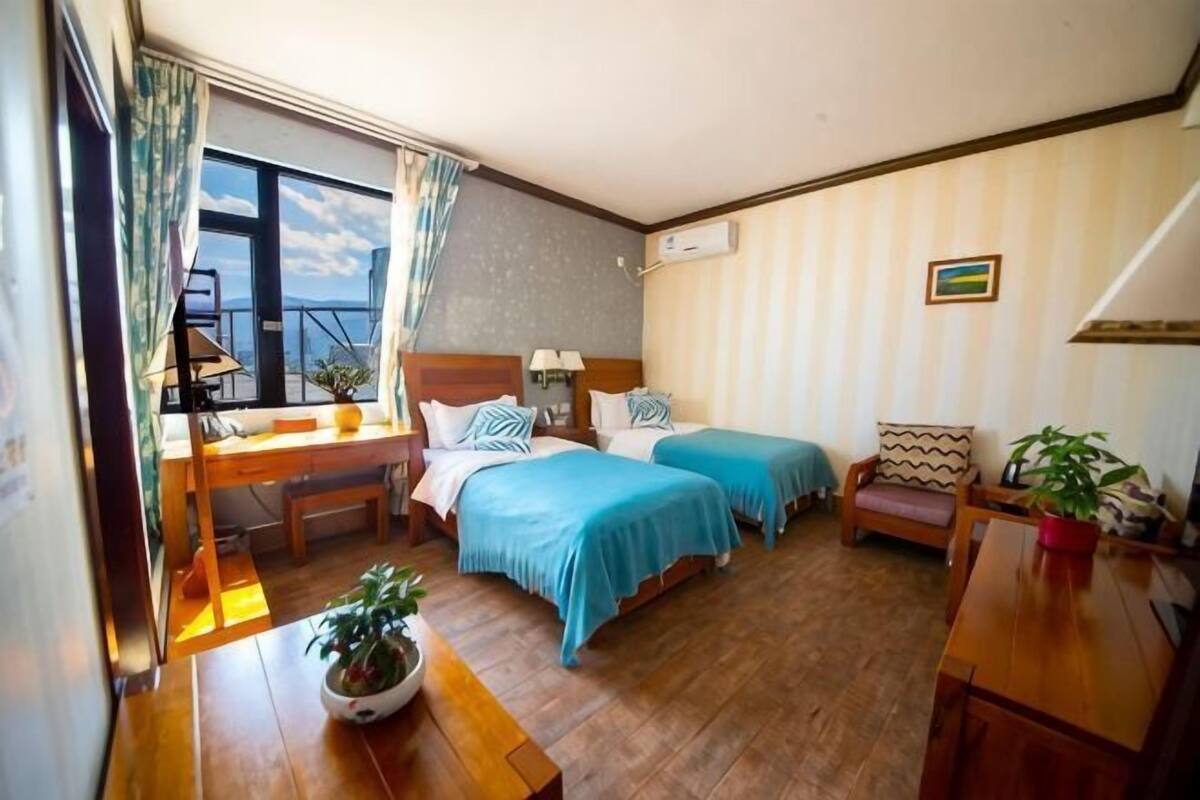 Quarto de hotel com duas camas, uma escrivaninha com luminária, uma cadeira, uma janela com cortinas e um vaso de plantas.