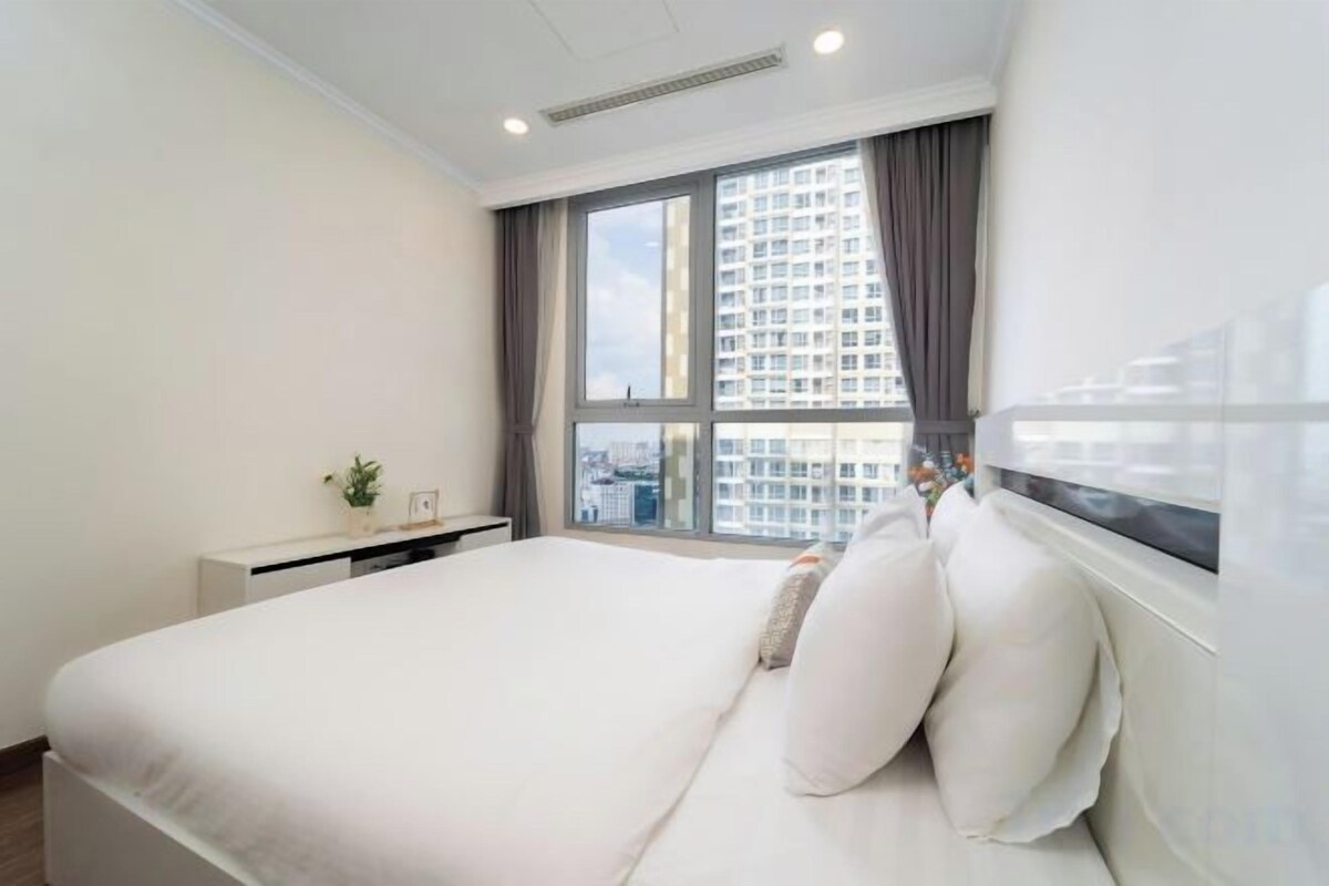 Quarto de hotel com uma cama grande, uma mesa de cabeceira, uma janela com vista para um prédio alto e paisagem urbana.