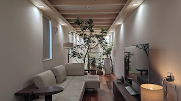 Uma sala de estar moderna com um sofá seccional, uma mesinha, uma luminária e uma televisão. Há plantas em vasos e teto de madeira.