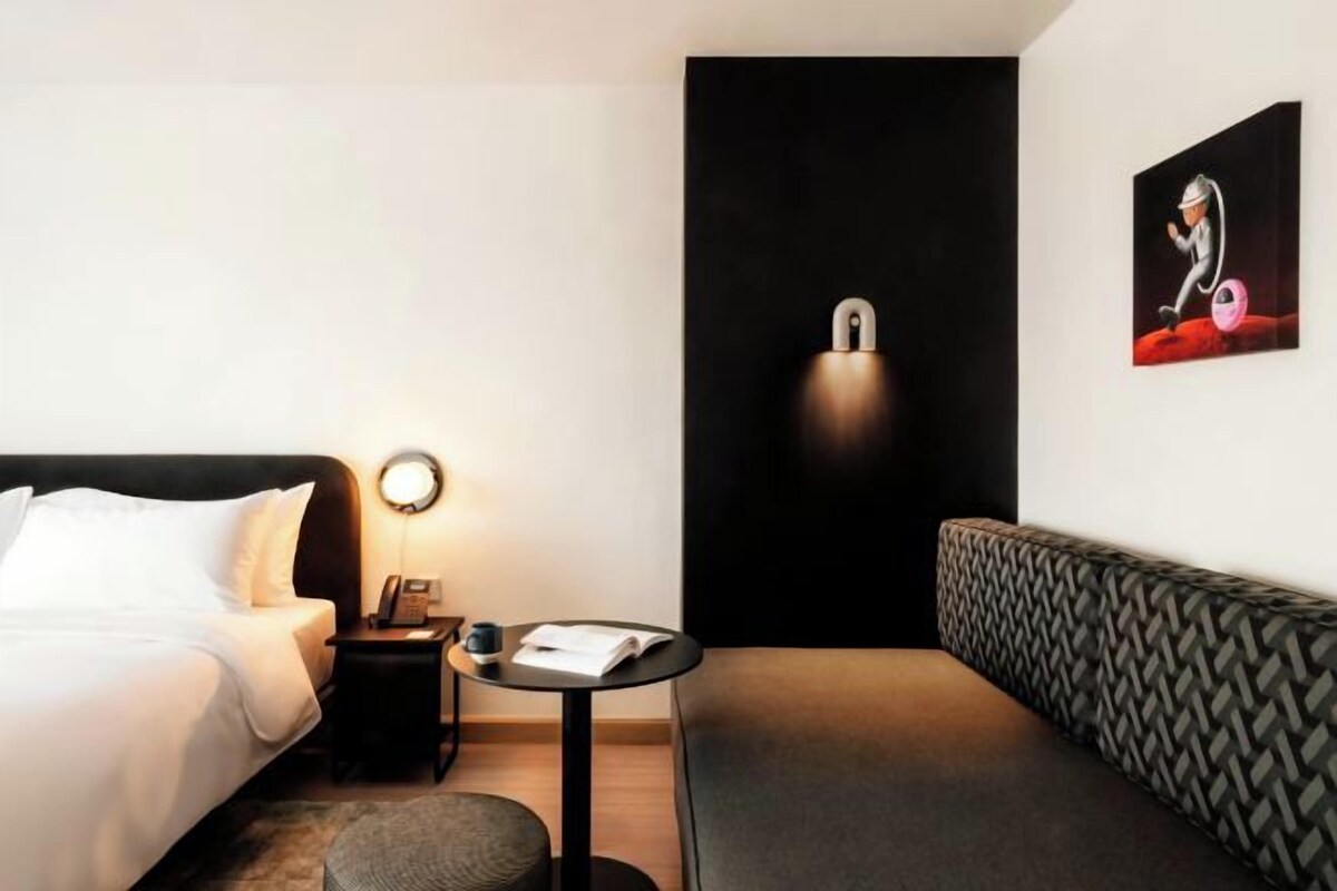 Quarto de hotel com cama, mesa de cabeceira, abajur e um sofá.