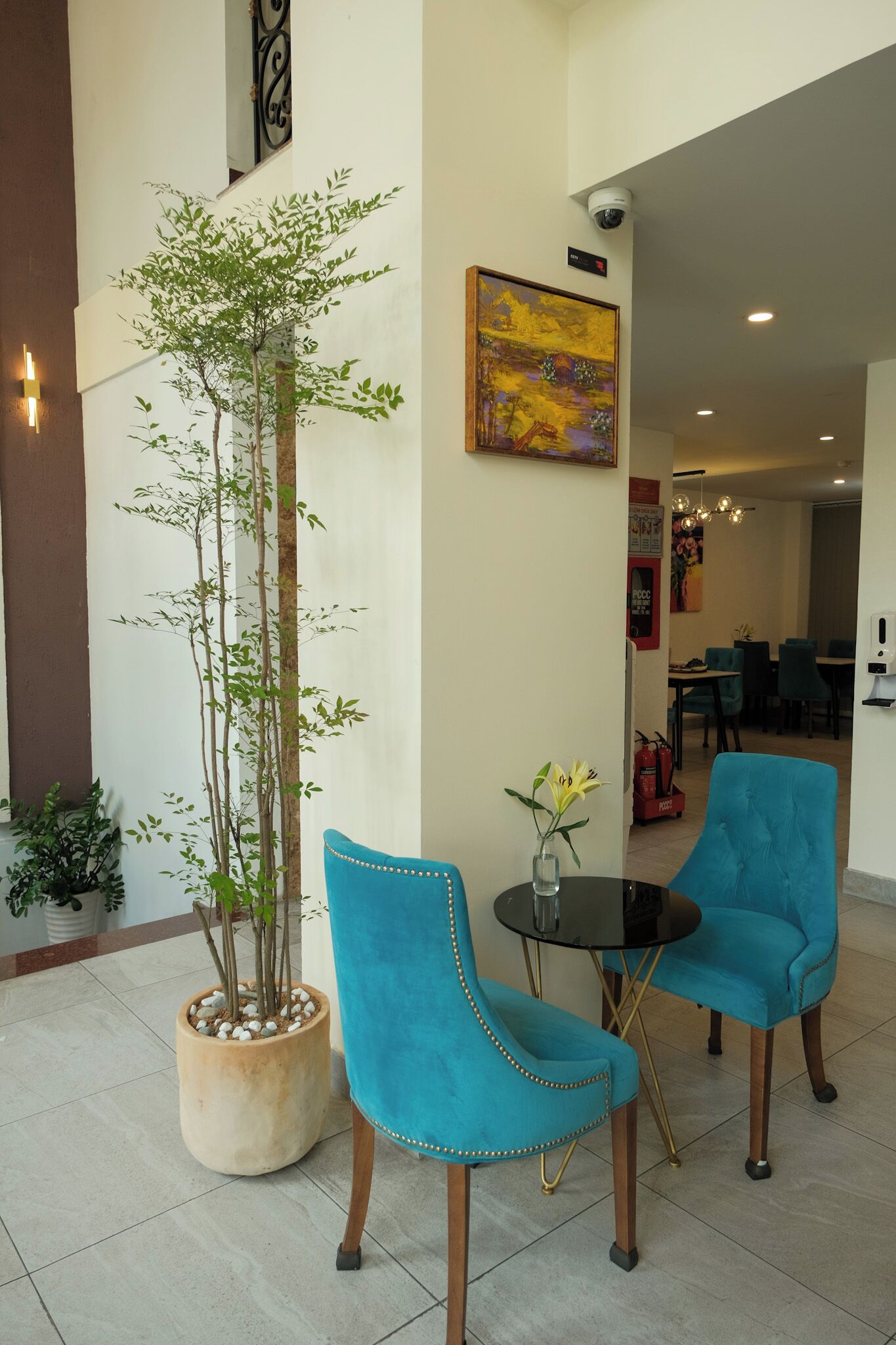 A lobby with two teal chairs, a small round table, a large potted plant, and a painting on the wall.