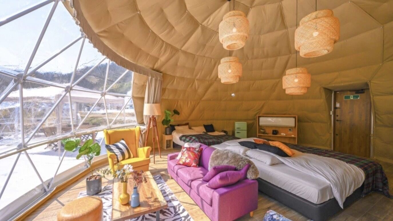 Acolhimento em tenda confortável, com cama, sofá e luminárias pendentes.