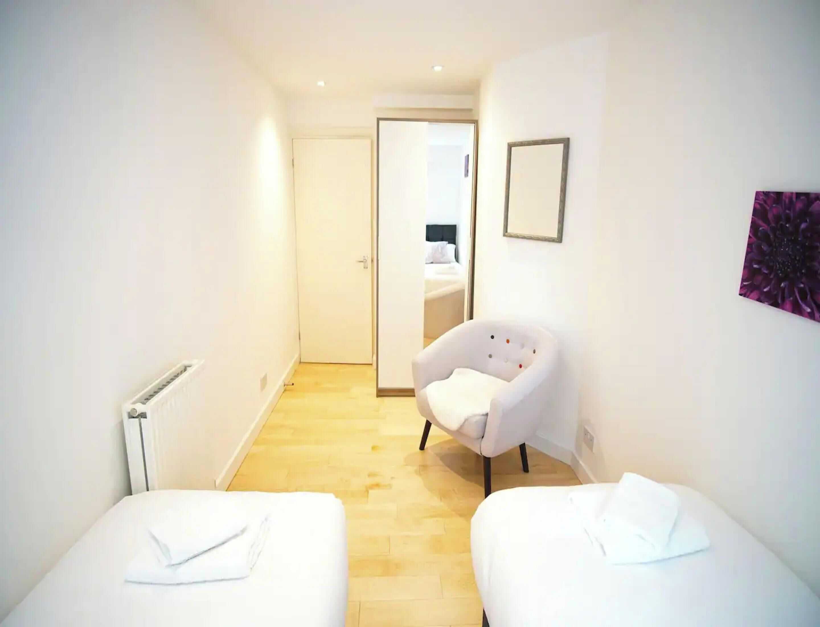 Quarto de hotel estreito com duas camas de solteiro, um radiador, uma cadeira branca e uma porta que dá para outro quarto.