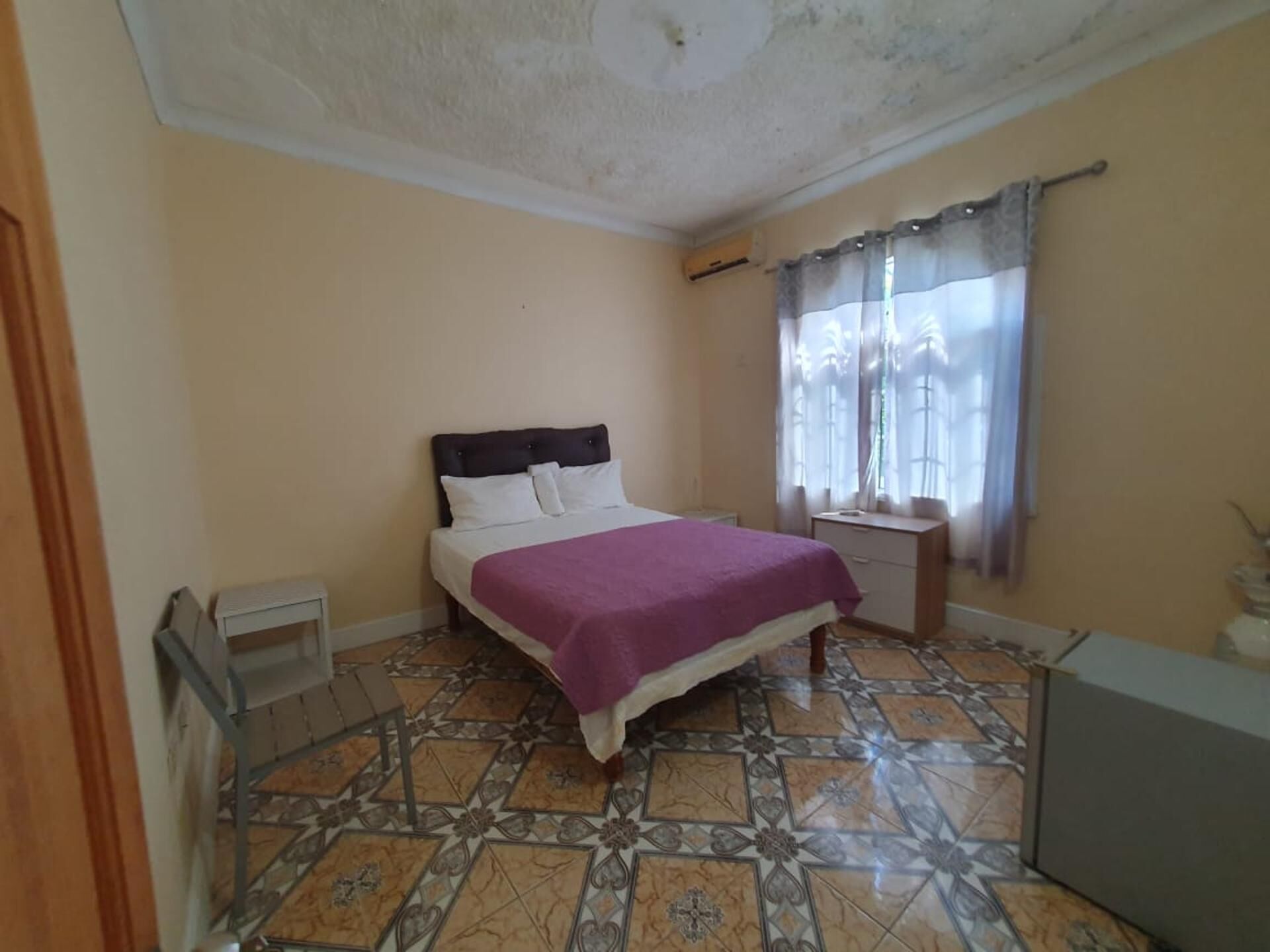 Quarto com cama, mesa de cabeceira, cômoda e janela com cortinas.