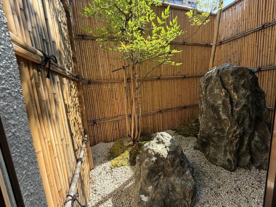 Uma cerca de bambu, uma árvore e pedras em um jardim.
