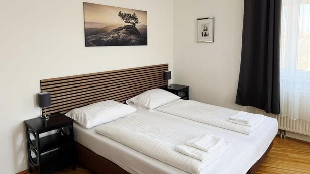 Uma cama de casal com roupa de cama branca, uma cabeceira de madeira, duas mesas de cabeceira com abajures, um quadro na parede e uma janela com cortinas.