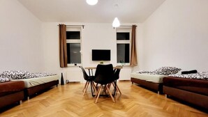 Quarto com soalho de madeira em padrão herringbone, dois sofás com almofadas estampadas, uma mesa de jantar com cadeiras, uma televisão de ecrã plano na parede e janelas com cortinas.