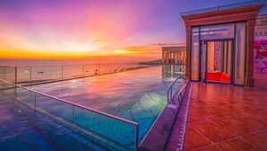 Piscina infinita com barreira de vidro, área de estar coberta e vista do pôr do sol sobre o oceano.