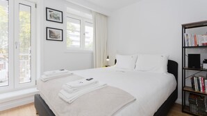 Cama bem arrumada com roupa de cama branca e toalha dobrada, uma estante ao lado com livros, e janela com cortinas.