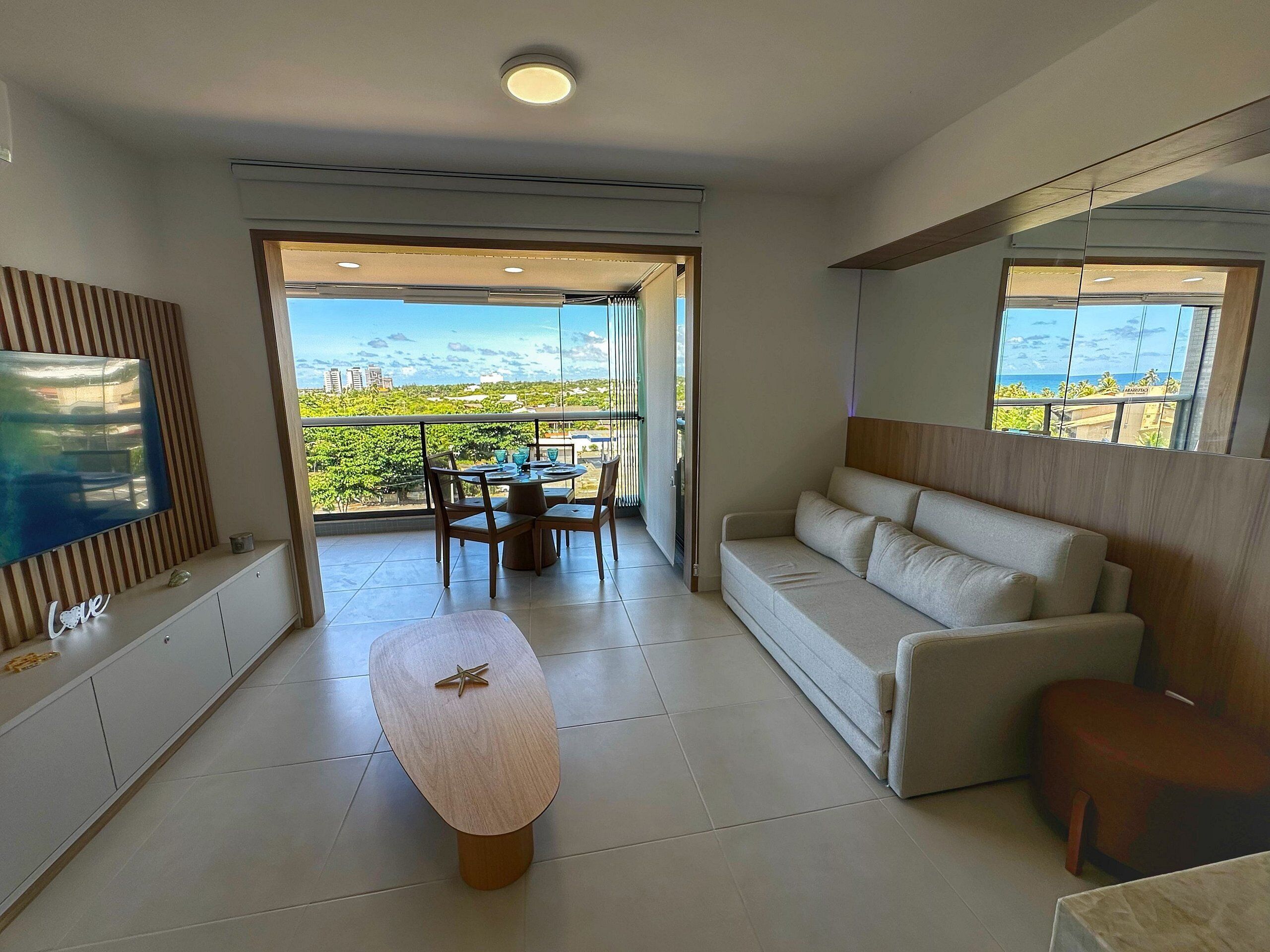 Uma sala de estar moderna com sofá, mesa de jantar e cadeiras, televisão na parede e uma varanda com vista.
